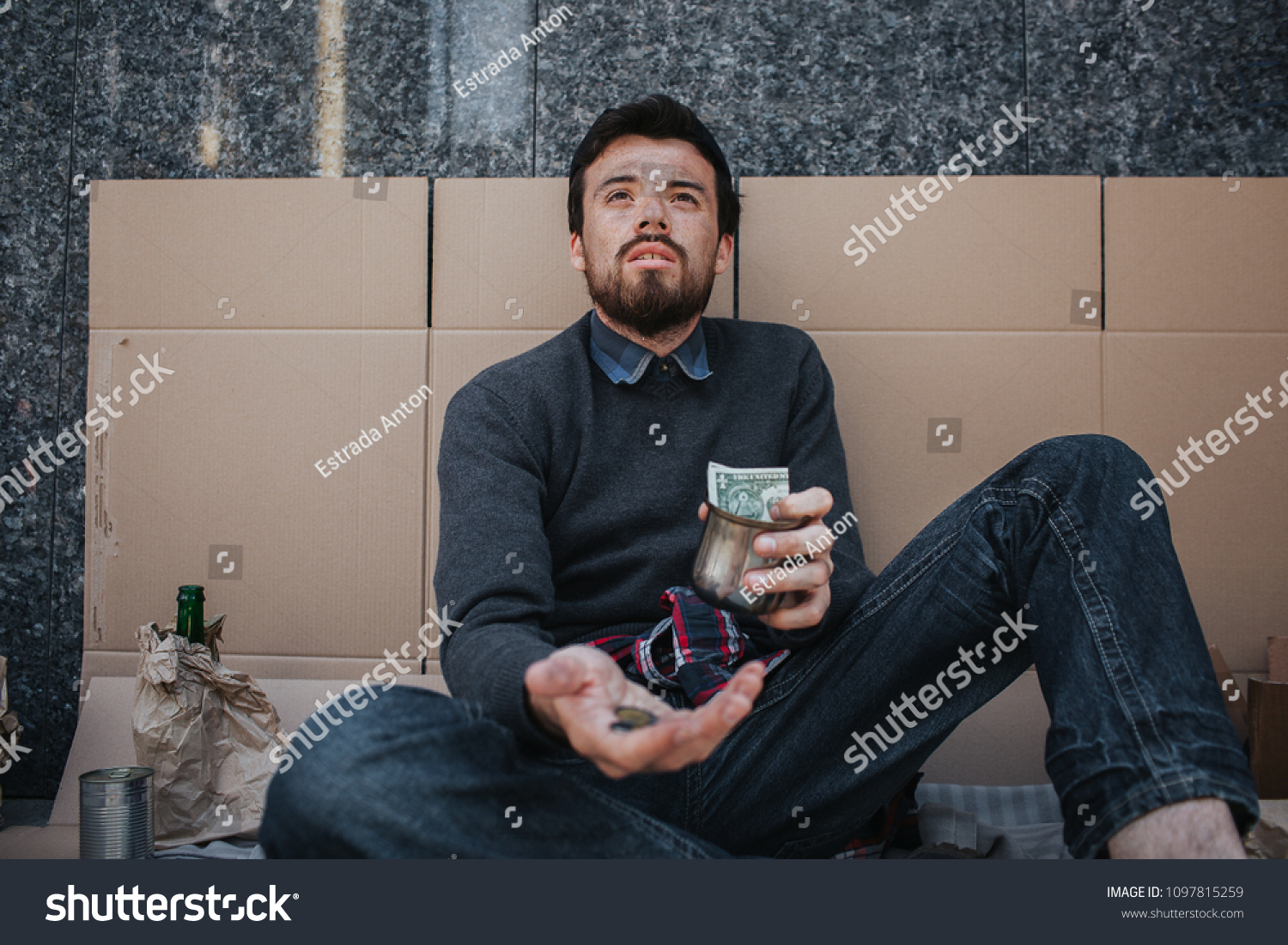 Adult beggar is sitting on cardboard and begging money. He is looking ...