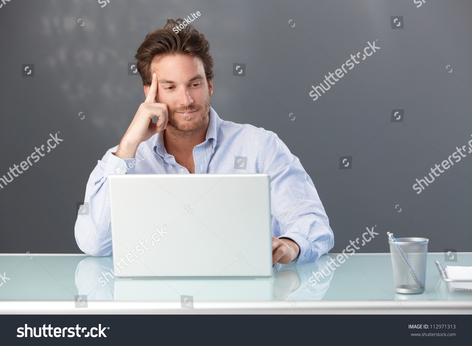Smiling handsome male office worker sitting at desk looking at laptop ...