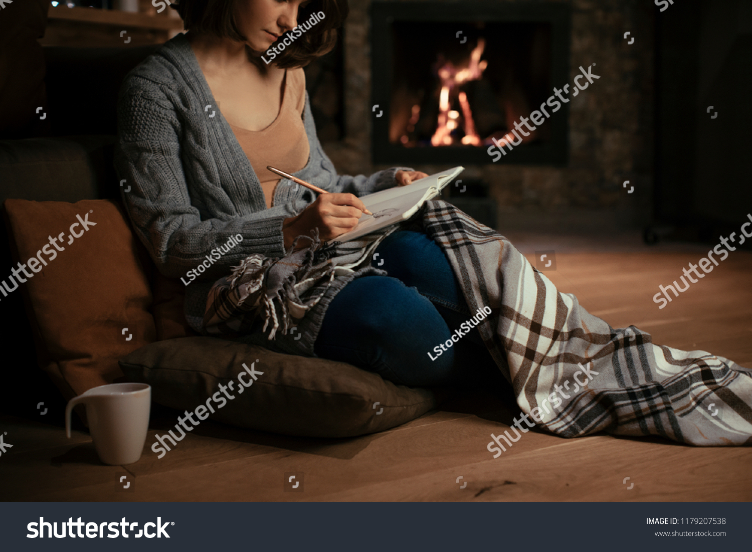 Beautiful woman sitting by the fireplace in cozy sweater and writing in ...