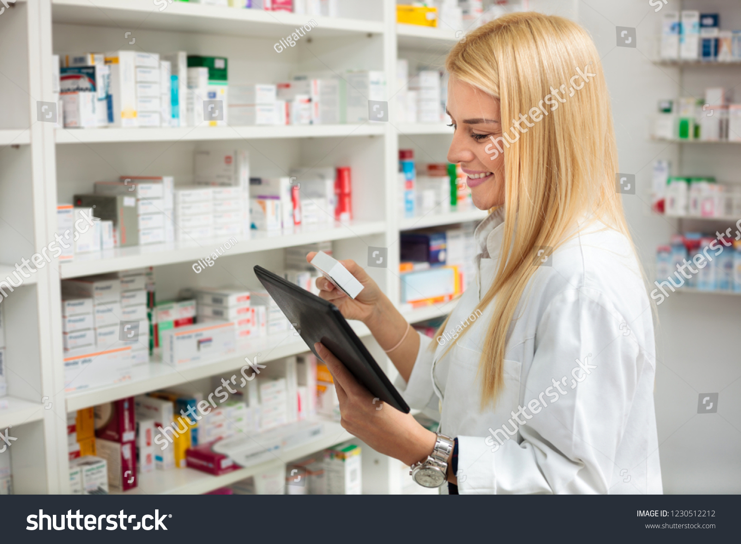 Beautiful smiling young female pharmacist standing in front of shelves with medications holding ...