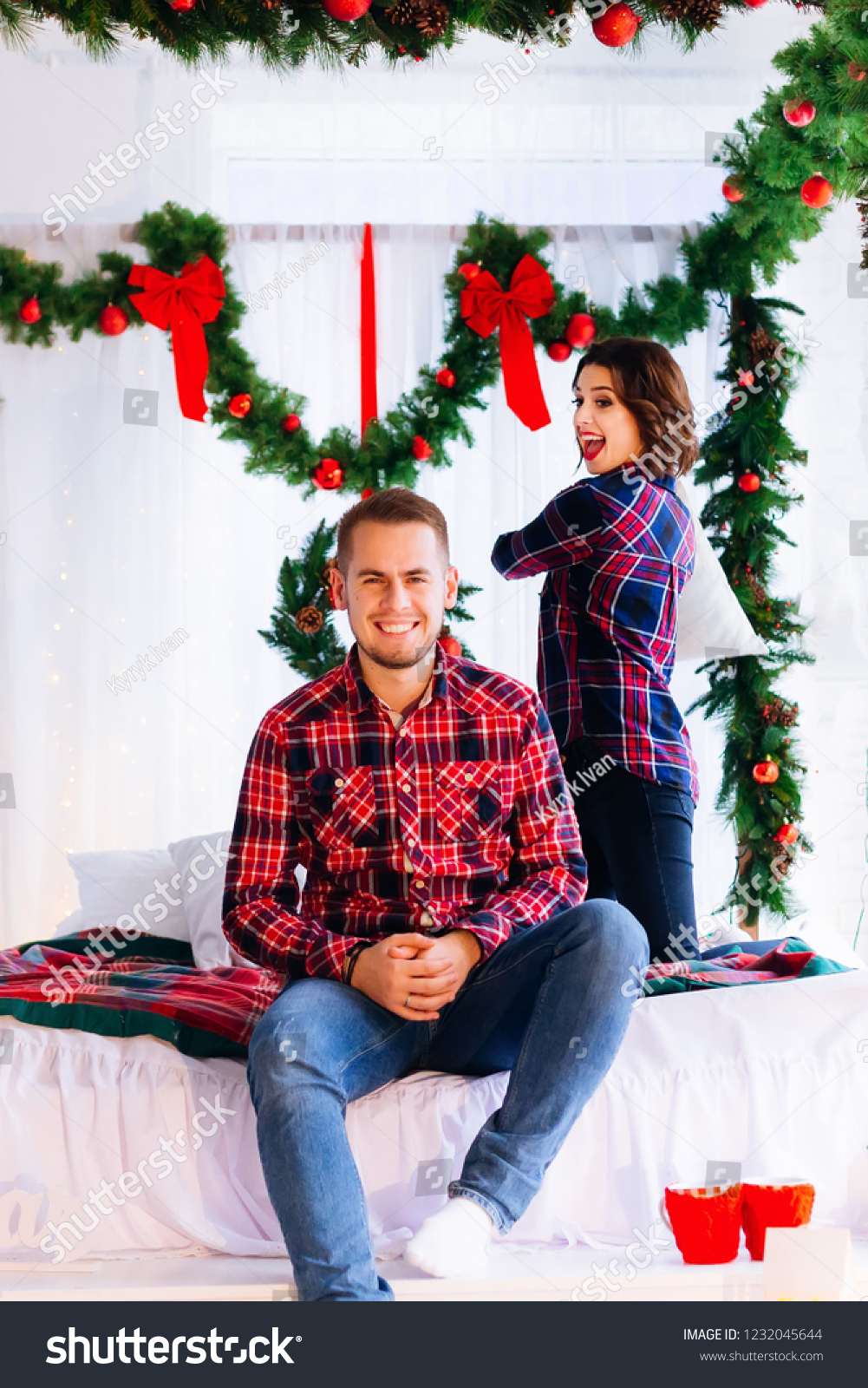 The guy is sitting on the bed and and smiles. the girl on the bed holds a pillow in hands and ...