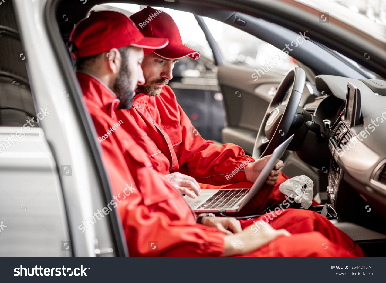 Two male auto mechanics in red uniform diagnosing car with computer ...