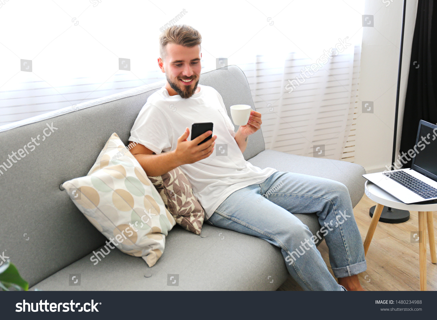 Bearded guy wearing blank white t-shirt and denim pants sitting alone at home on grey textile ...