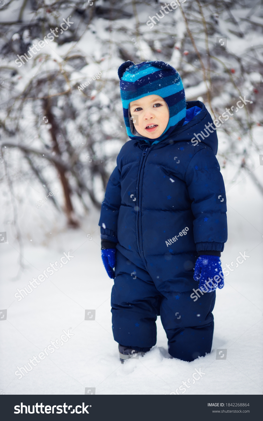 little boy in snowsuit and funny hat walking on snow in winter outdoors ...