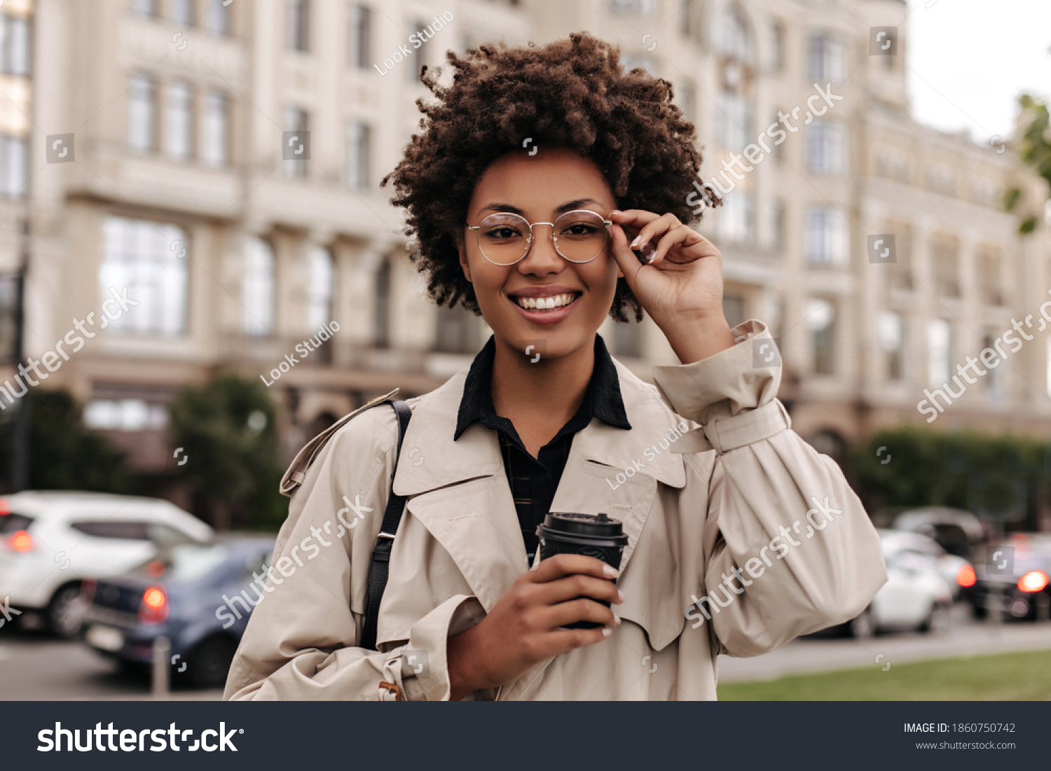 Cheerful happy brunette curly dark-skinned woman in beige trench coat puts on round eyeglasses ...