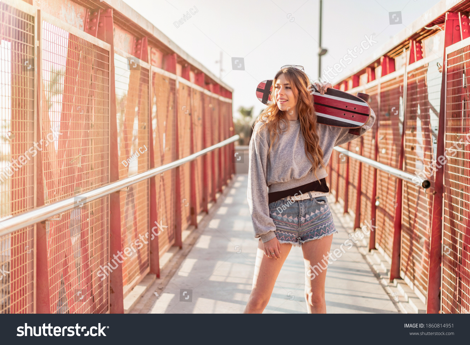 Skater woman standing at a red bridge holding a skateboard. Cool urban outfit california style ...