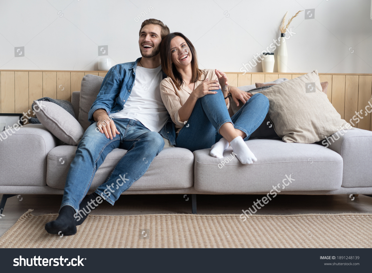 Happy young Caucasian couple relax on cozy sofa at home hugging look in distance dreaming ...