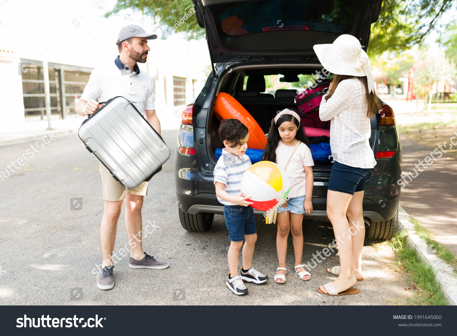 Hispanic parents packing their suitcases in the trunk of their car with their children. Young ...