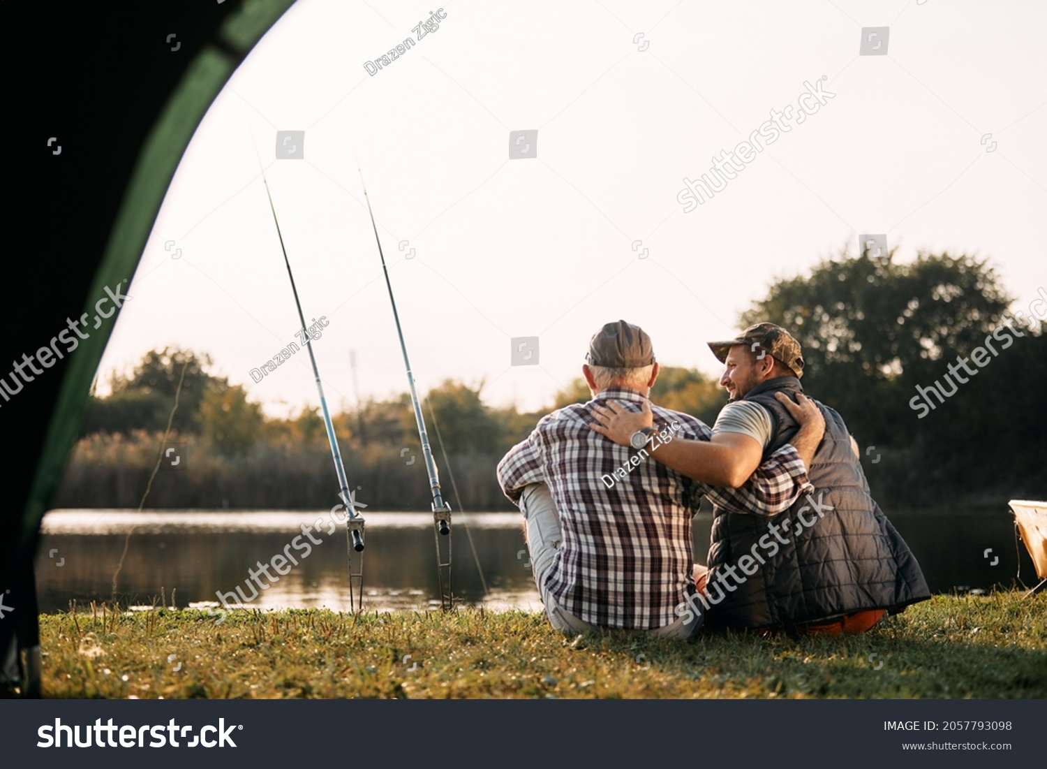 Back view of happy man embracing his senior father while fishing during their camping day. Copy ...