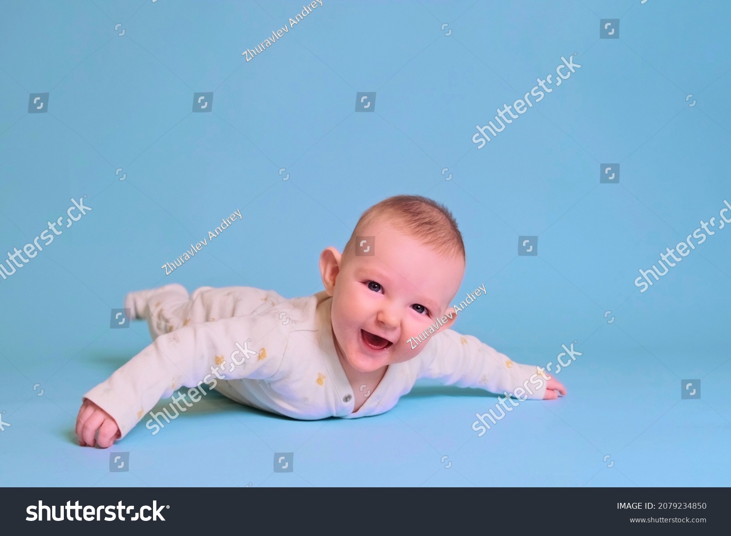 Happy infant baby boy laughing lying on his tummy blue studio background. Four month old child ...