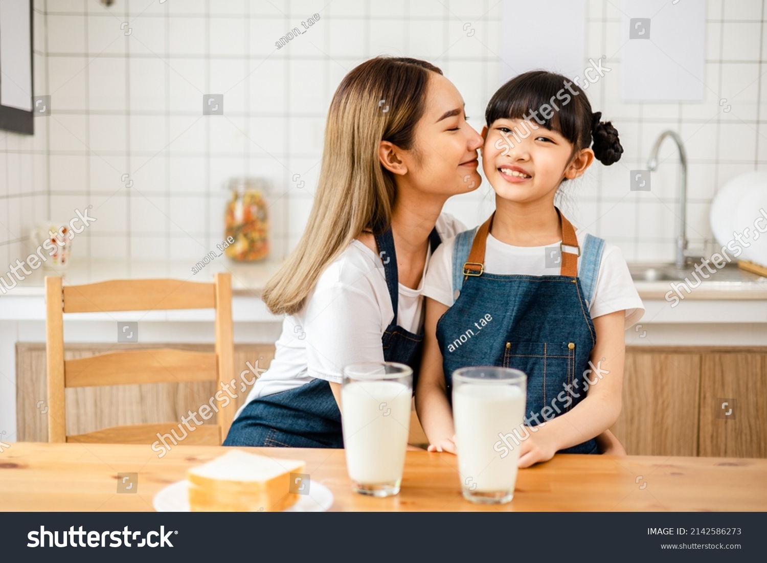 Asian teen girl drinking milk with her mom and eating bread sitting at kitchen table.Loving mom ...