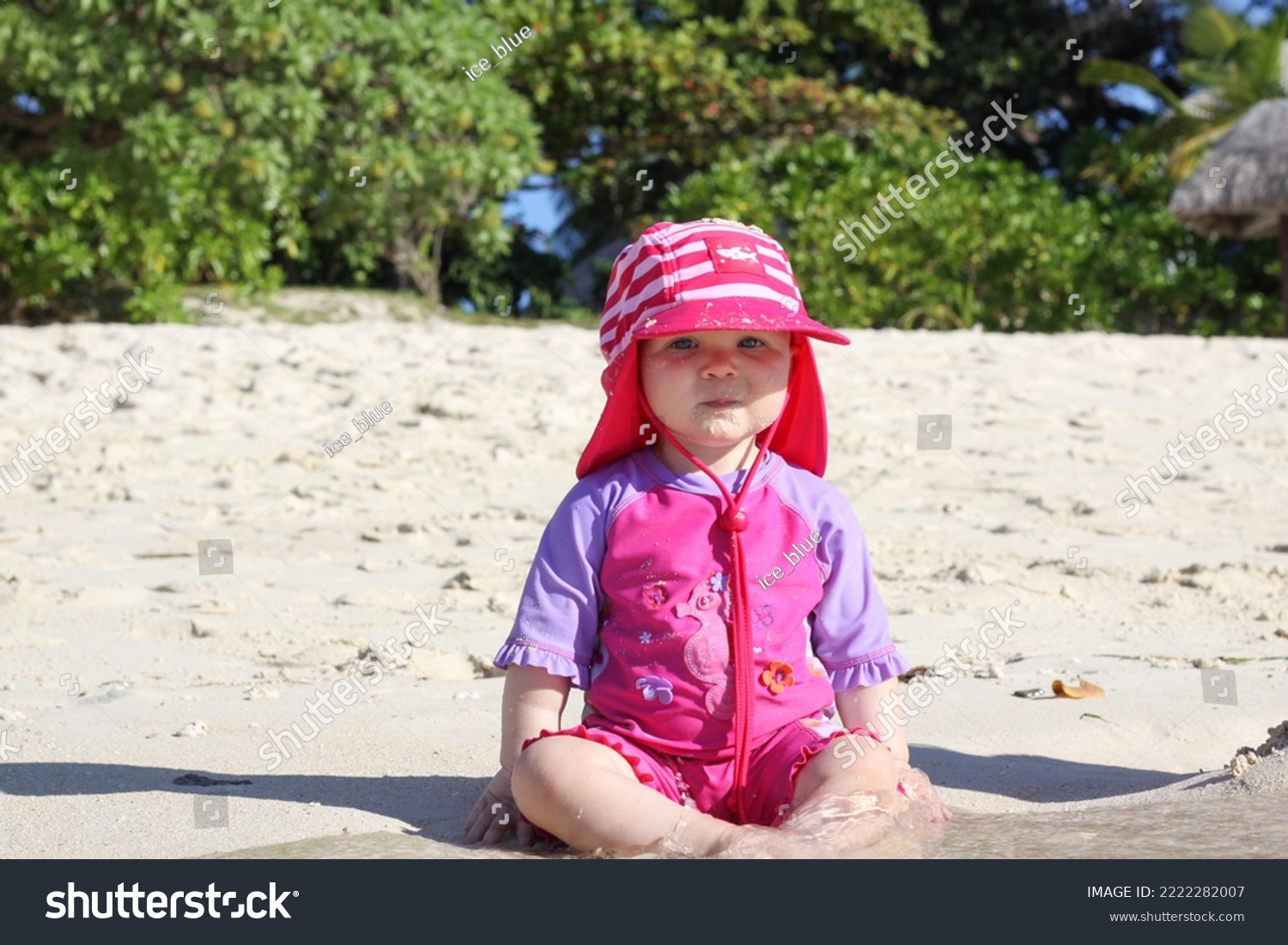 A 10 month old baby girl sitting on the sand in the shallow water at a ...