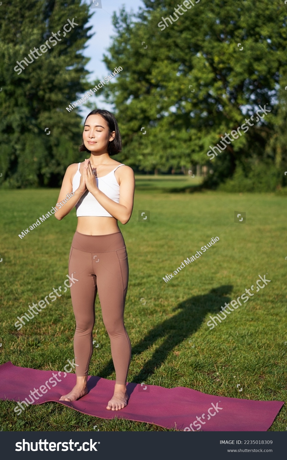 Vertical shot of asian woman standing in asana doing yoga exercises on fresh air in park wearing ...