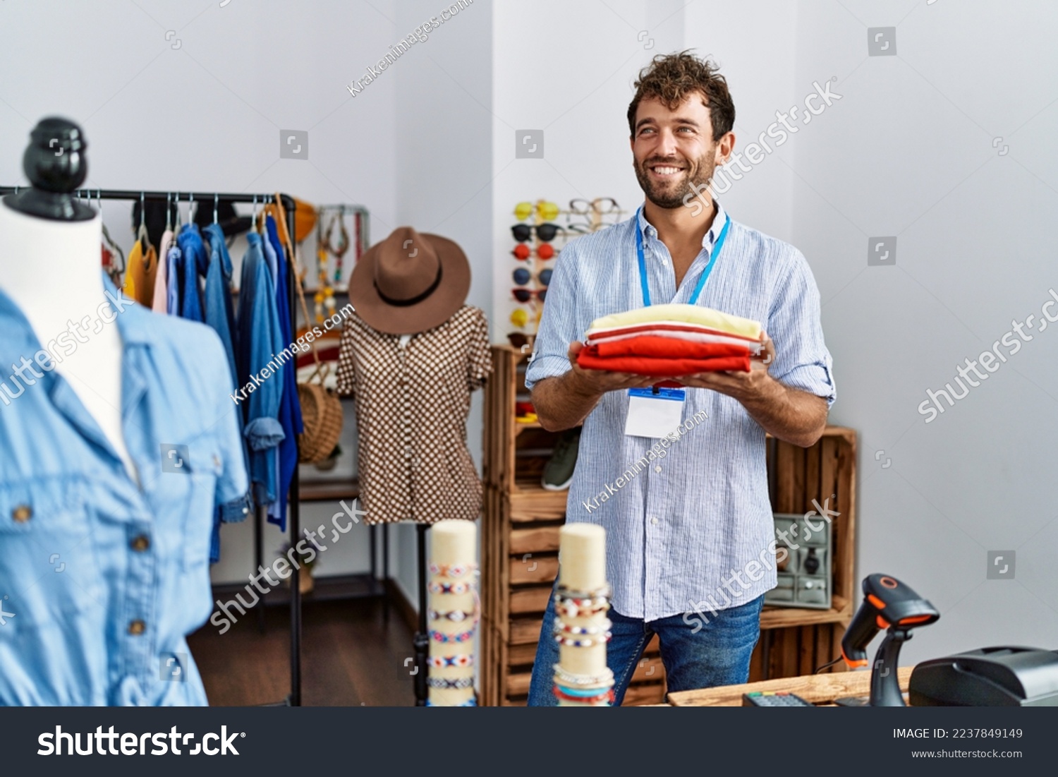 Young hispanic shopkeeper man smiling happy holding stack of sweater at ...