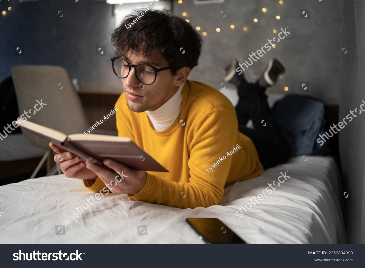 Student lying on a bed and studying with focus and concentration in college dormitory. Young man ...