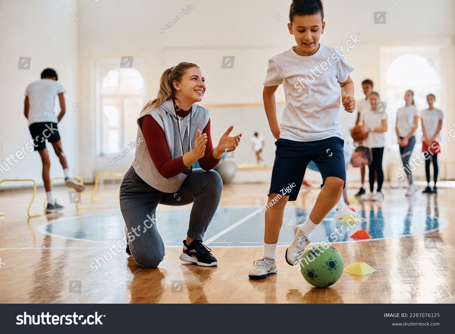 Schoolboy leading soccer ball among cones while sports teacher is ...