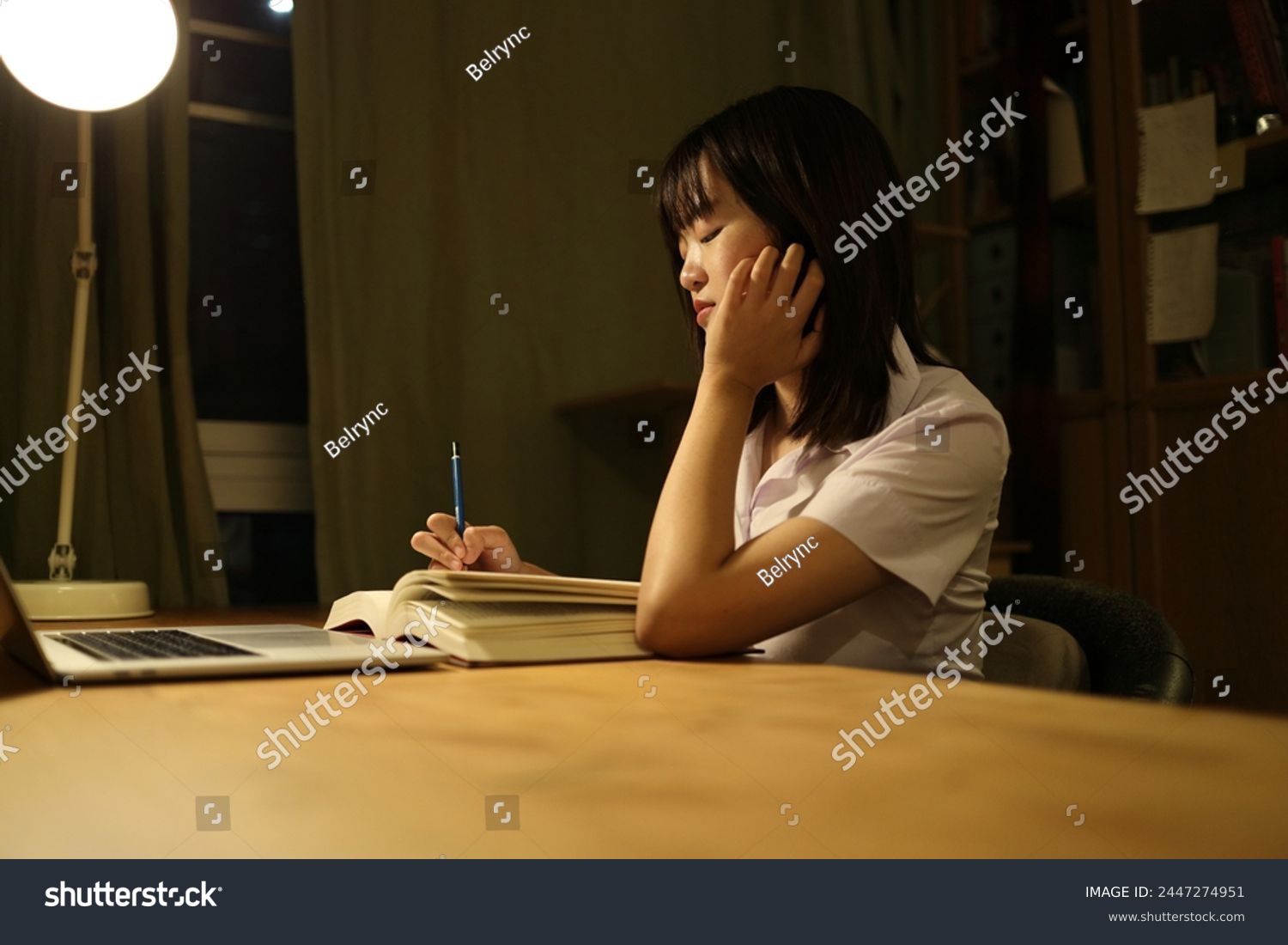 A young female student in a Thai university uniform sits at a desk in study doing homework and ...