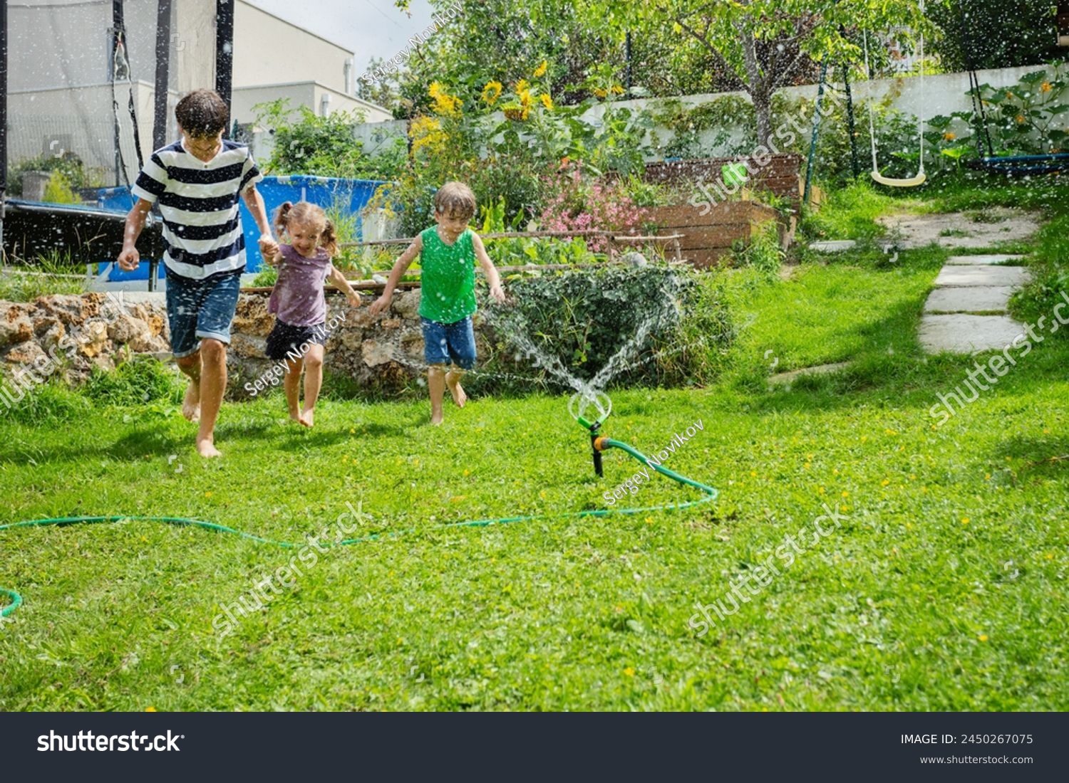 Two children and an teenager enjoy a carefree summer day sprinting through sprinkler water in a ...