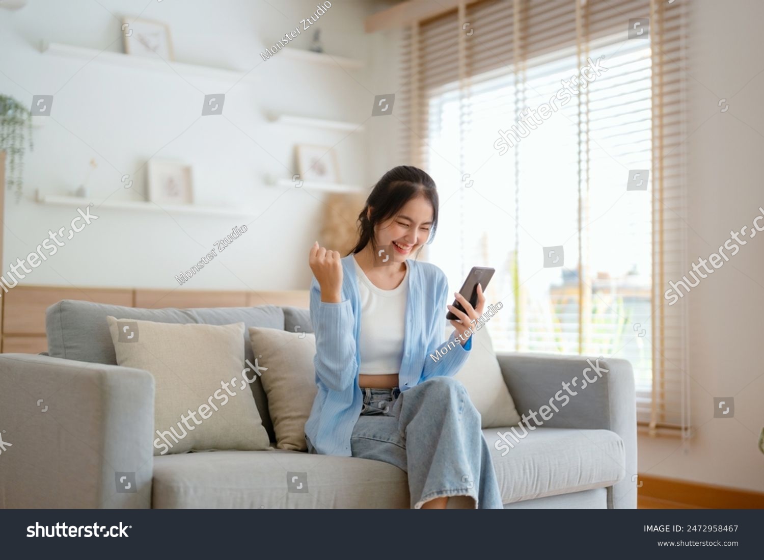 Smiling pretty young Asian woman sitting on cozy couch using funny mobile apps in living room ...
