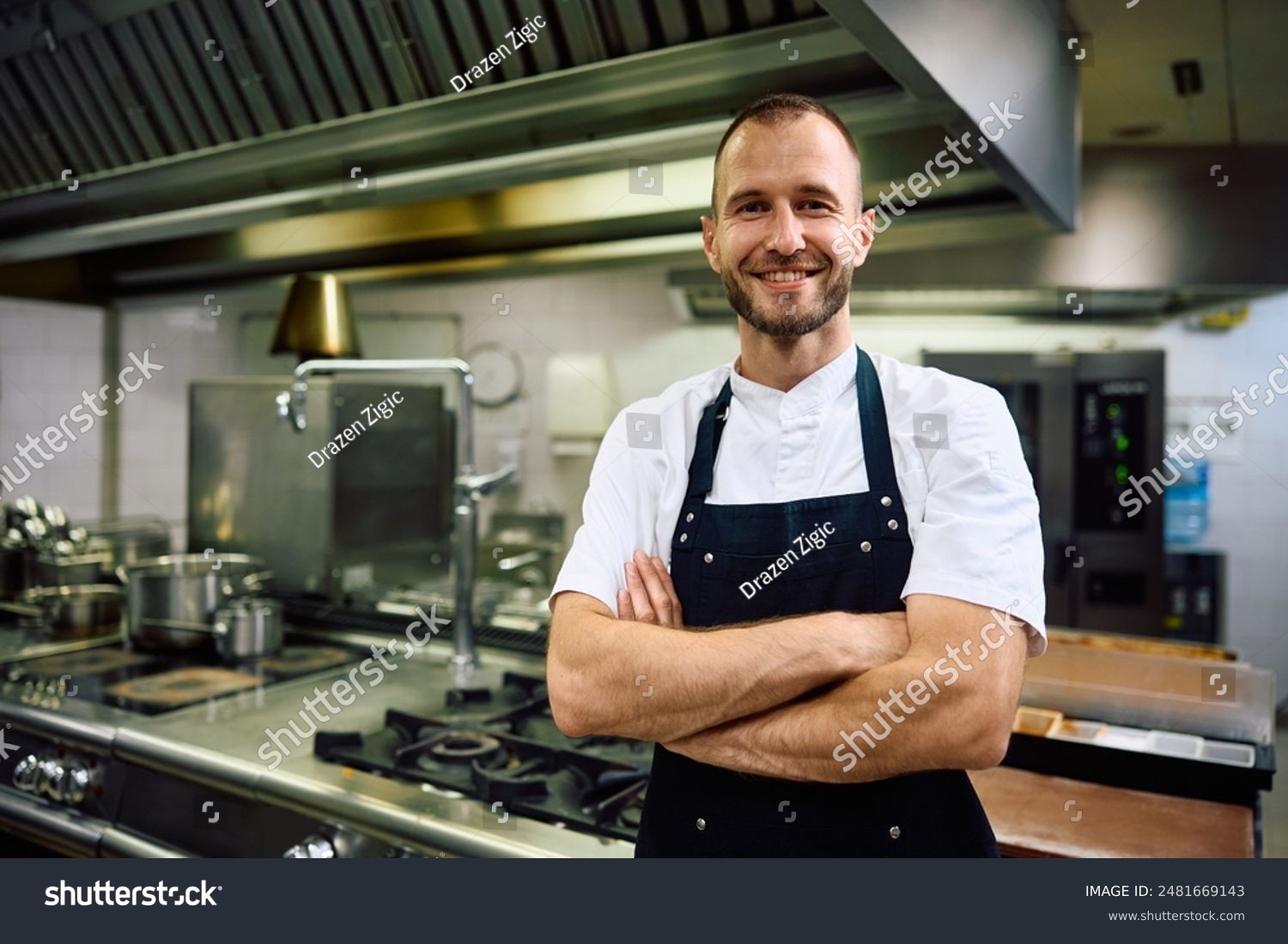 Confident chef standing with crossed arms in the kitchen at restaurant and looking at camera. _站 ...