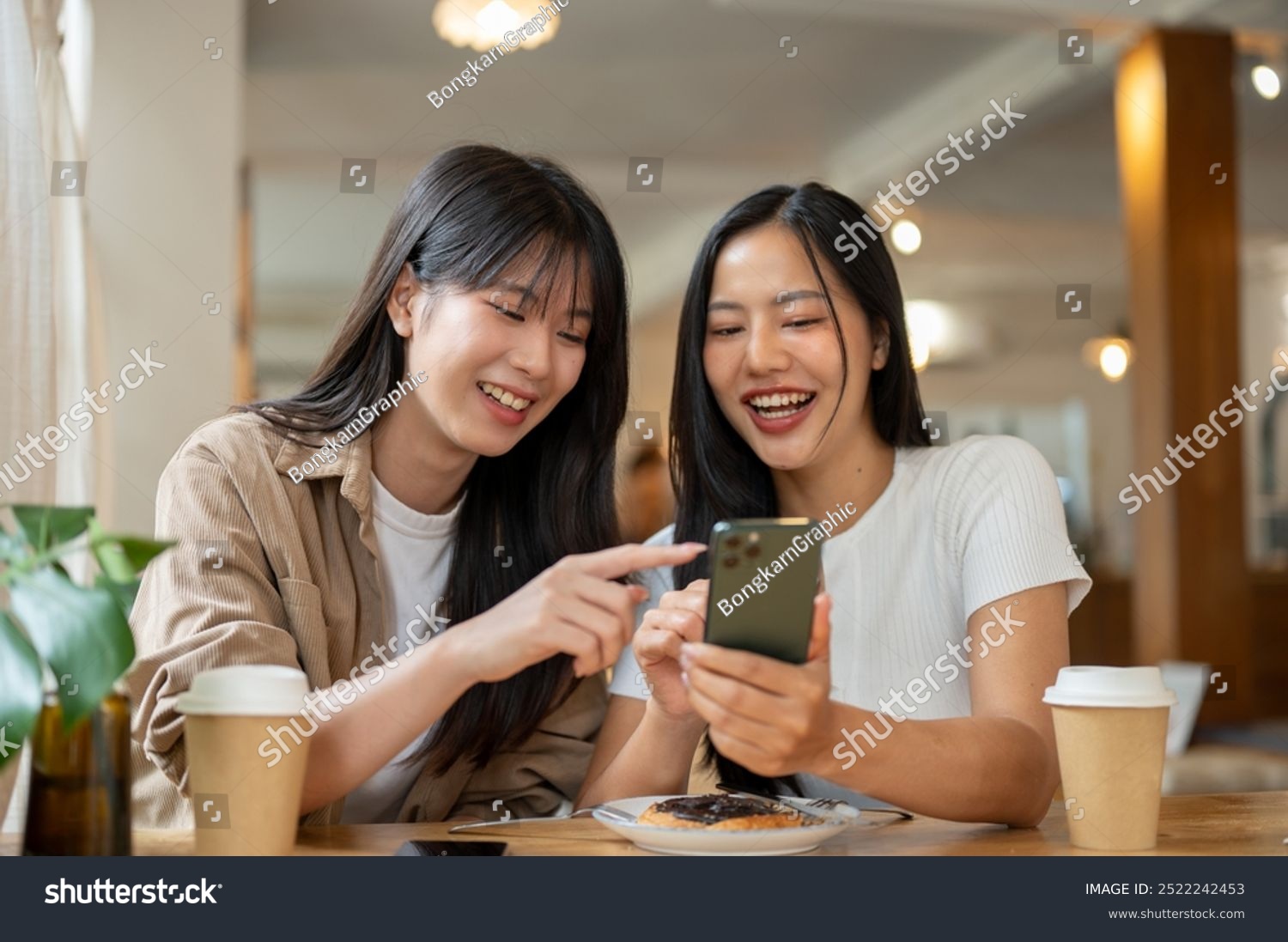 Two positive and charming Asian female friends are looking at something on a smartphone laughing ...