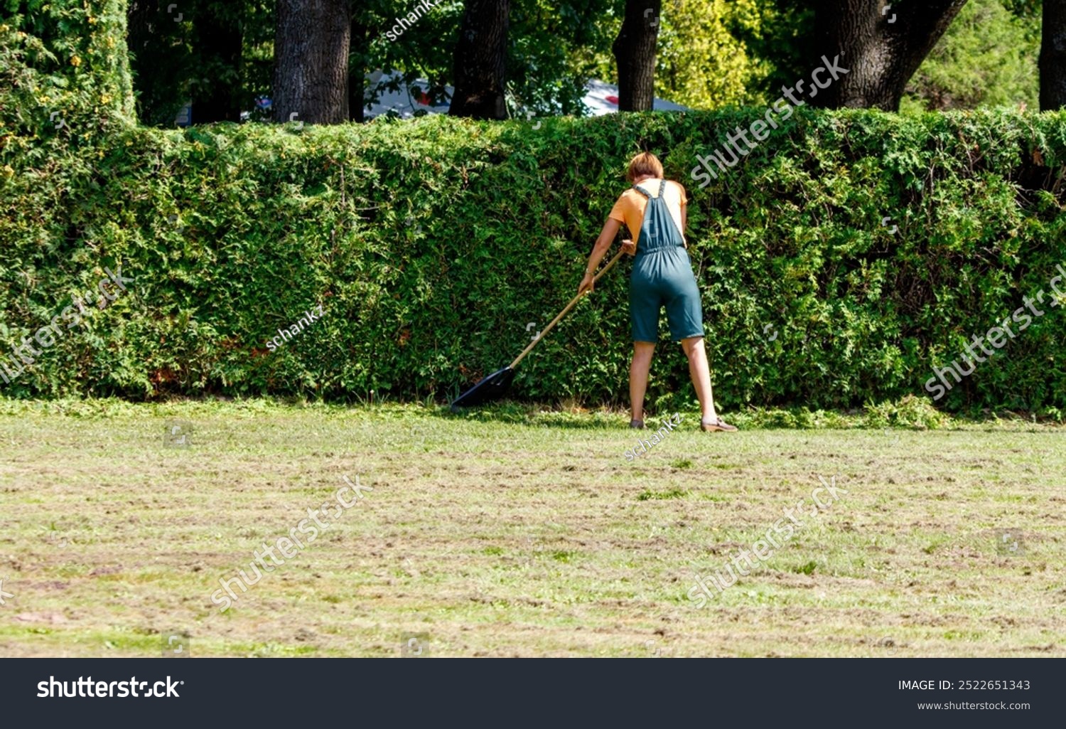 A woman in a green apron is walking through a hedge with a rake in her ...