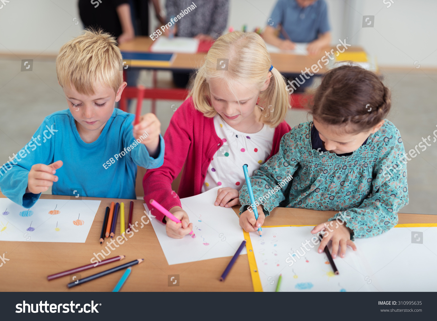 Three young children in kindergarten art class sitting at a desk side ...